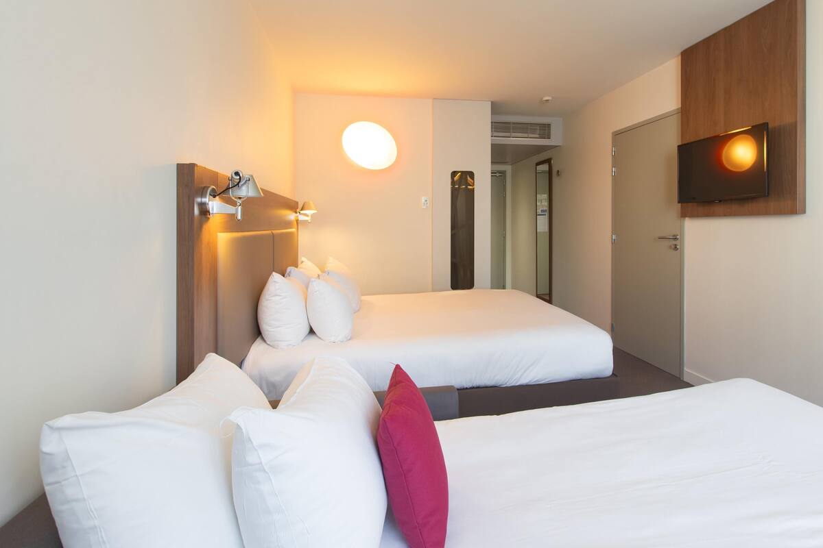 Quarto de hotel com duas camas de solteiro, uma cabeceira de madeira, um espelho e uma televisão presa à parede.