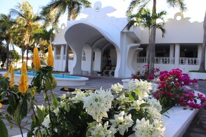 Um edifício com fachada branca e arcos, rodeado por plantas e flores tropicais.