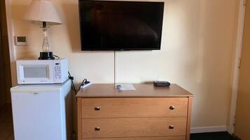 Quarto com televisão de tela plana na parede, micro-ondas sobre uma mesinha e cômoda de madeira com gavetas.