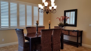 Uma sala de jantar com mesa, cadeiras, lustre, mesa consola com espelho e janela com persianas.