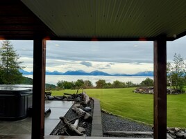 Vista de uma área externa coberta com terraço, hidromassagem e um campo gramado que se estende até a água, com montanhas ao longe.