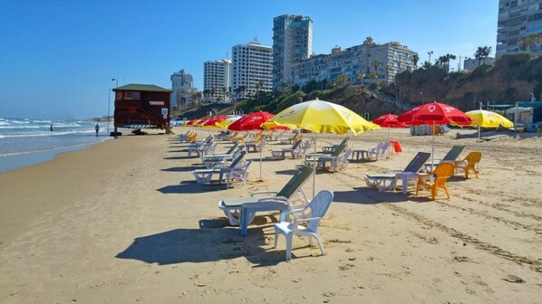 Uma praia com fileiras de espreguiçadeiras e guarda-sóis, uma torre de salva-vidas e edifícios altos ao fundo.