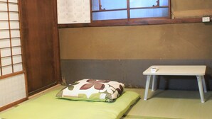 Quarto japonês tradicional com tatames, duas camas baixas com roupa de cama estampada com flores, um pequeno banco de madeira e uma janela com vista para uma paisagem distante.