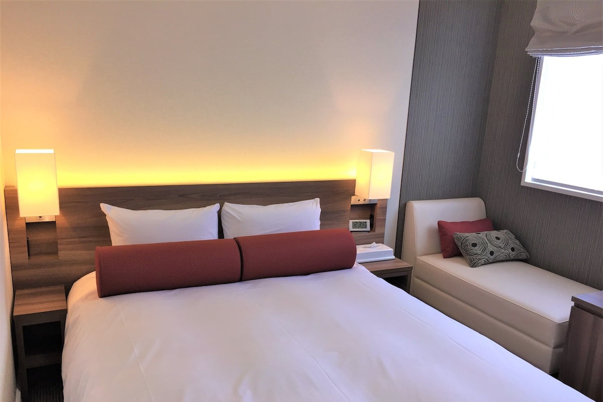 Quarto de hotel com cama grande, duas mesinhas de cabeceira, um criado-mudo, um sofá e uma janela com cortinas.