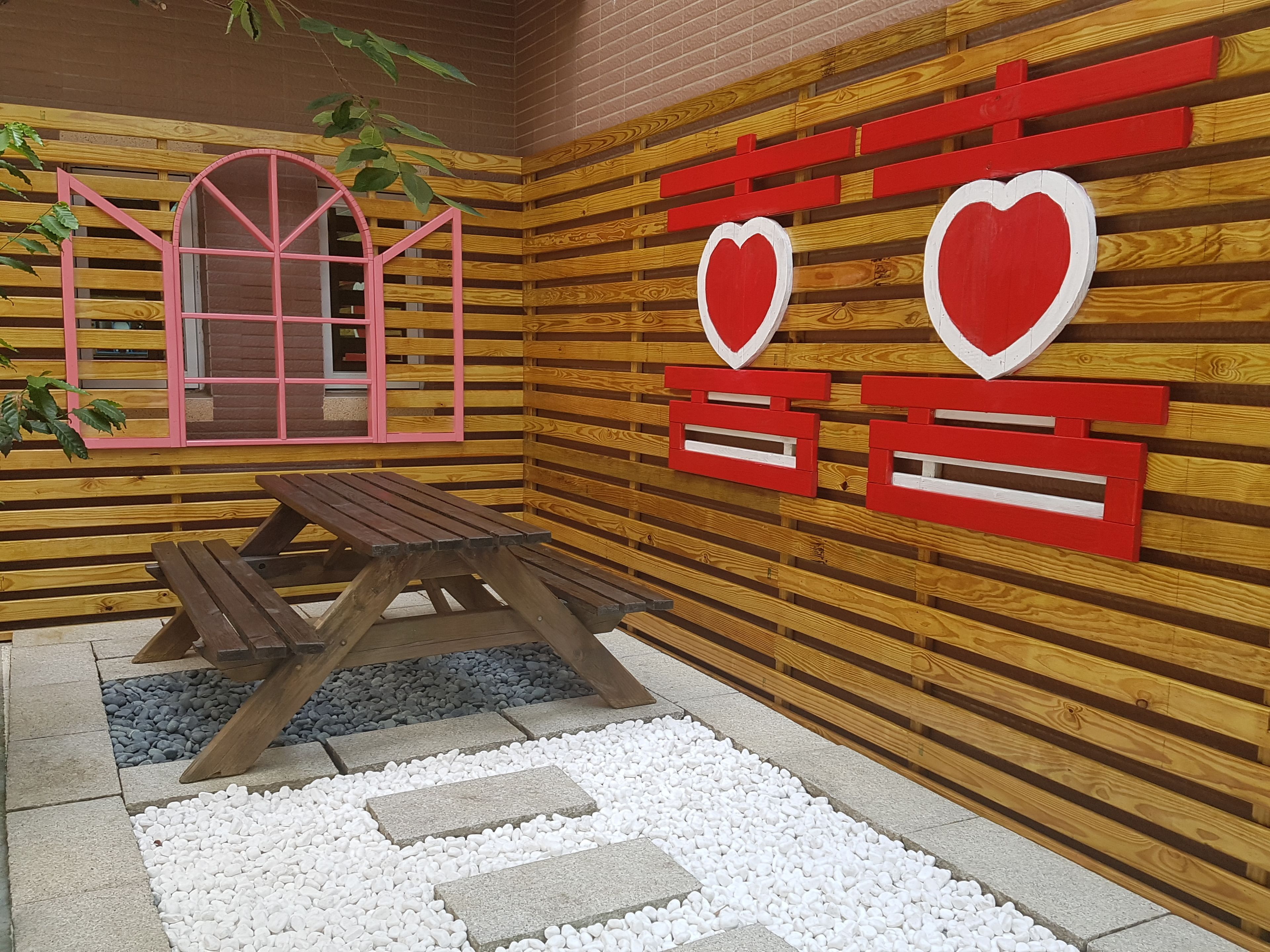Uma cabana de madeira com uma mesa de piquenique, uma caixa de correio vermelha e uma janela com moldura rosa.