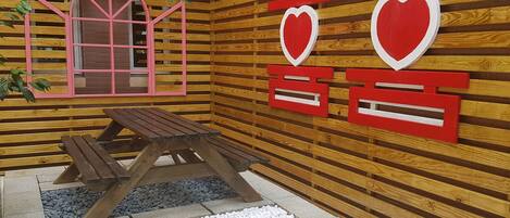 Uma cabana de madeira com uma mesa de piquenique, uma caixa de correio vermelha e uma janela com moldura rosa.