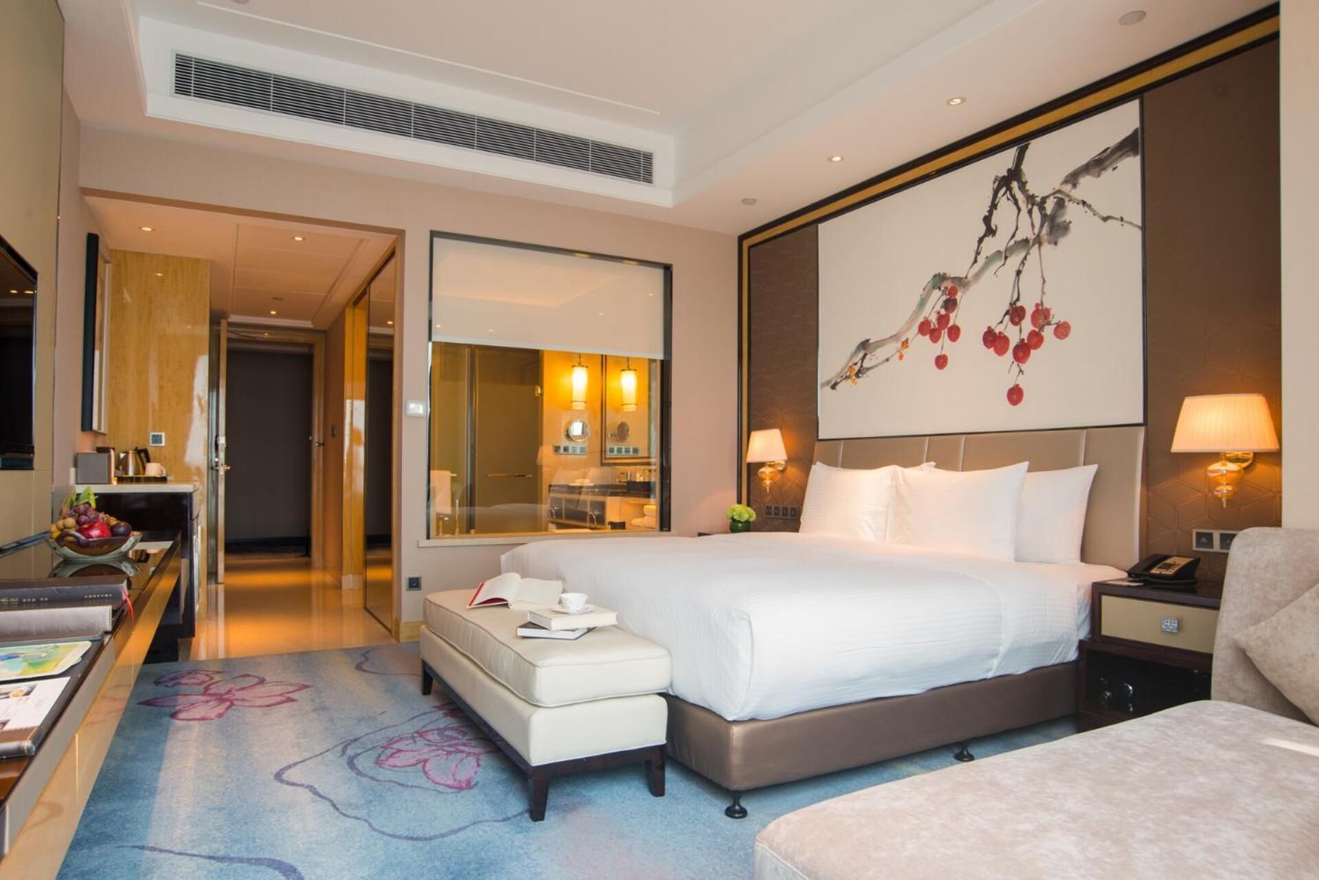 Quarto de hotel com uma cama grande, uma escrivaninha, uma cadeira, uma mesa pequena com uma tigela de frutas e um espelho amplo.