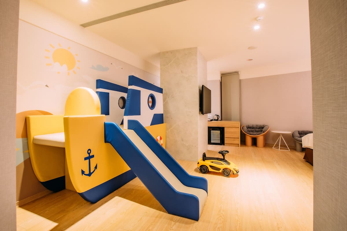 Espaço infantil com escorrega e assentos, um pequeno carro de brinquedo e uma televisão sobre suporte.