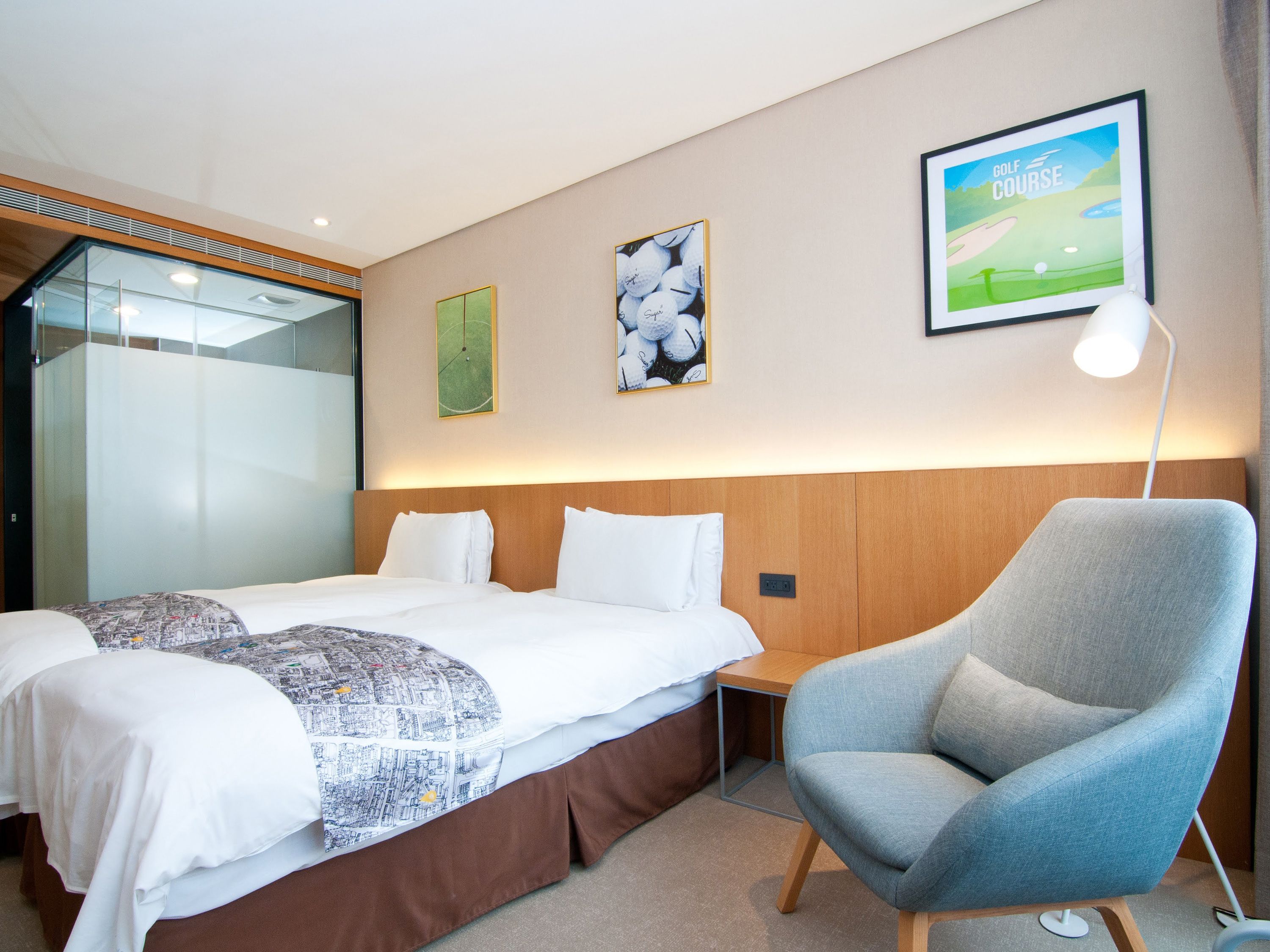 Quarto de hotel com cama, poltrona, escrivaninha e televisão na parede exibindo um campo de golfe.