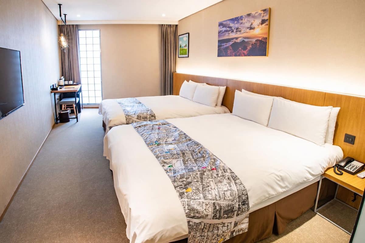 Quarto de hotel com duas camas, televisão, secretária e telefone, e um quadro na parede.