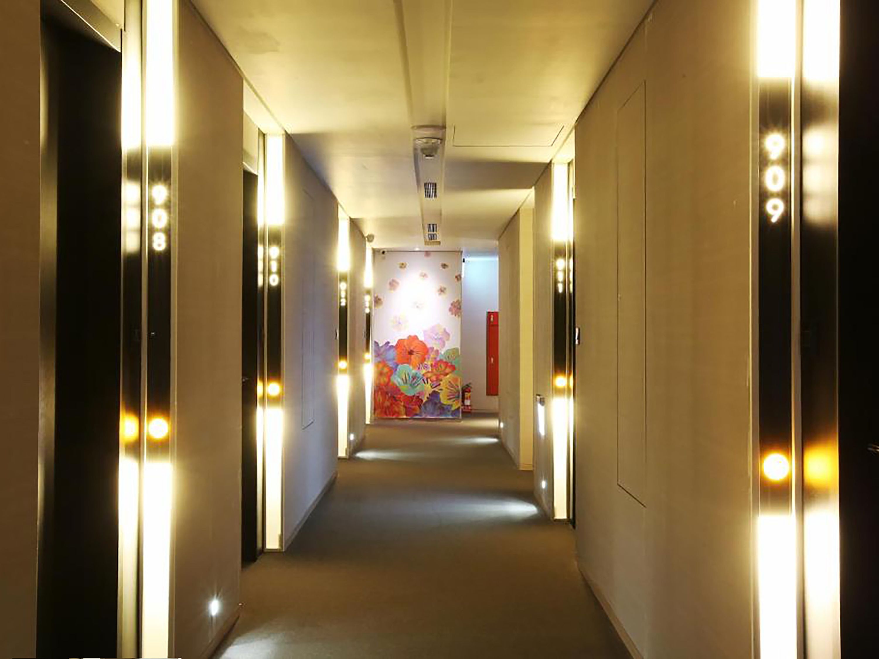 Um corredor de hotel com piso carpete, paredes com luminárias e um mural floral colorido no final.