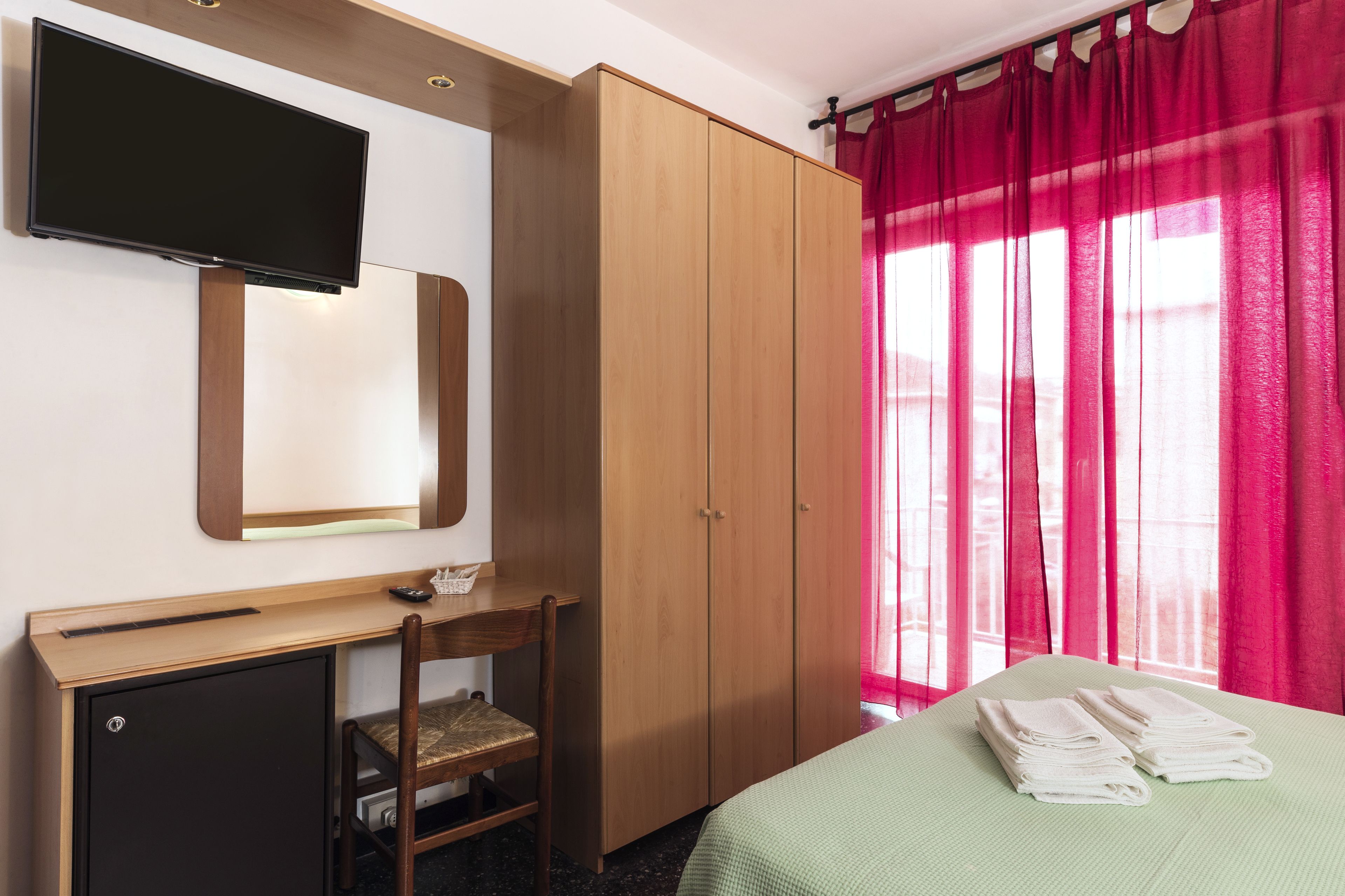 Quarto de hotel com cama, escrivaninha, cadeira, guarda-roupa, televisão e janela com cortinas.