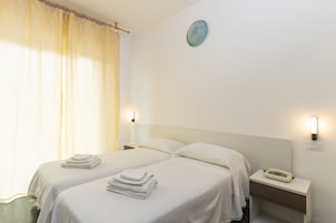 Quarto de hotel com duas camas, uma mesinha de cabeceira com telefone e um relógio na parede.