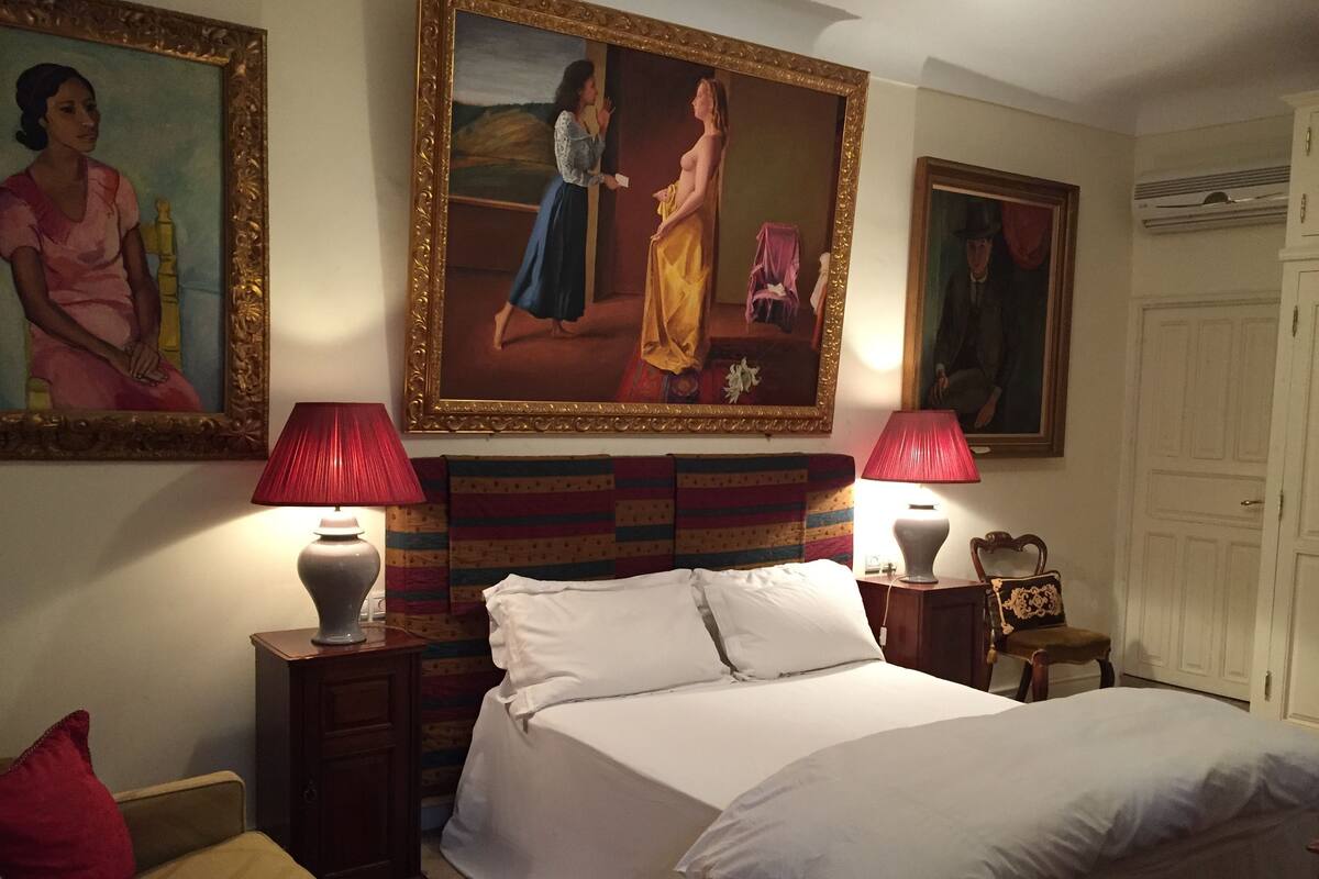 Quarto de hotel com uma cama grande, duas mesas de cabeceira com luminárias, uma cabeceira colorida e três quadros nas paredes.