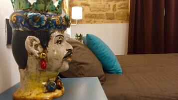 Uma escultura de cabeça em cerâmica com motivos florais sobre uma mesa, num quarto com parede de pedra, cama com roupa de cama castanha e almofada azul.