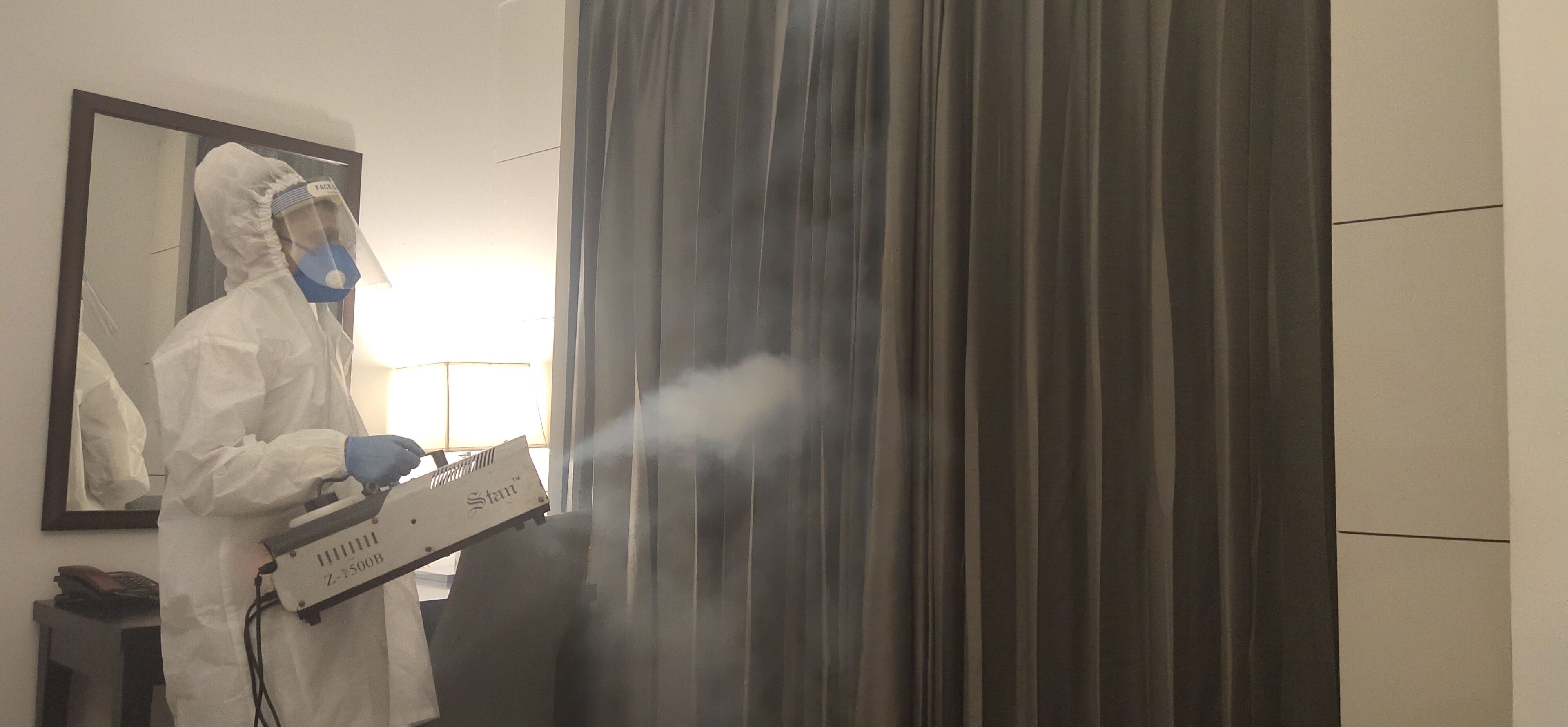 Uma pessoa vestindo um traje de proteção utilizando um pulverizador em um quarto com cortina.