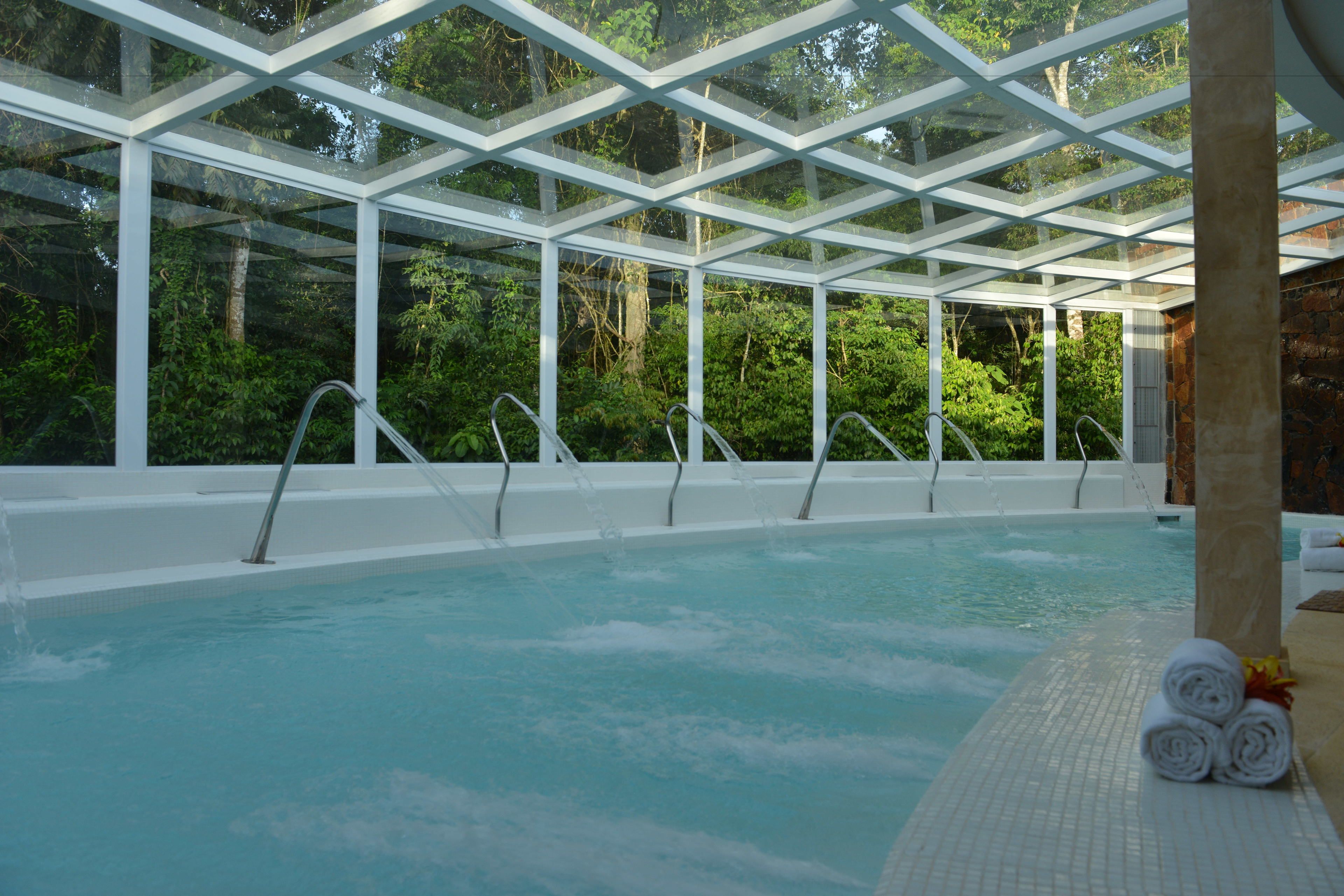 Piscina interior com paredes de vidro transparente, rodeada por vegetação exuberante.