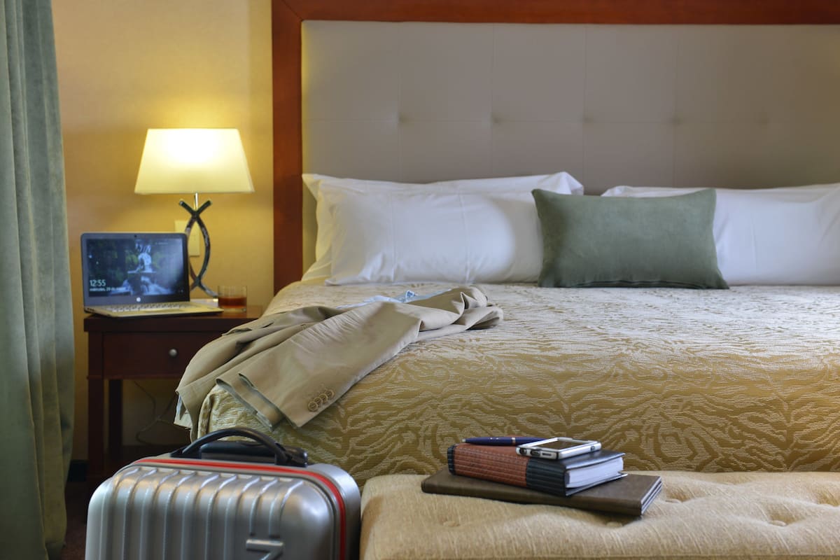 Quarto de hotel com cama, mesa de cabeceira, abajur, laptop, mala e uma pilha de livros.
