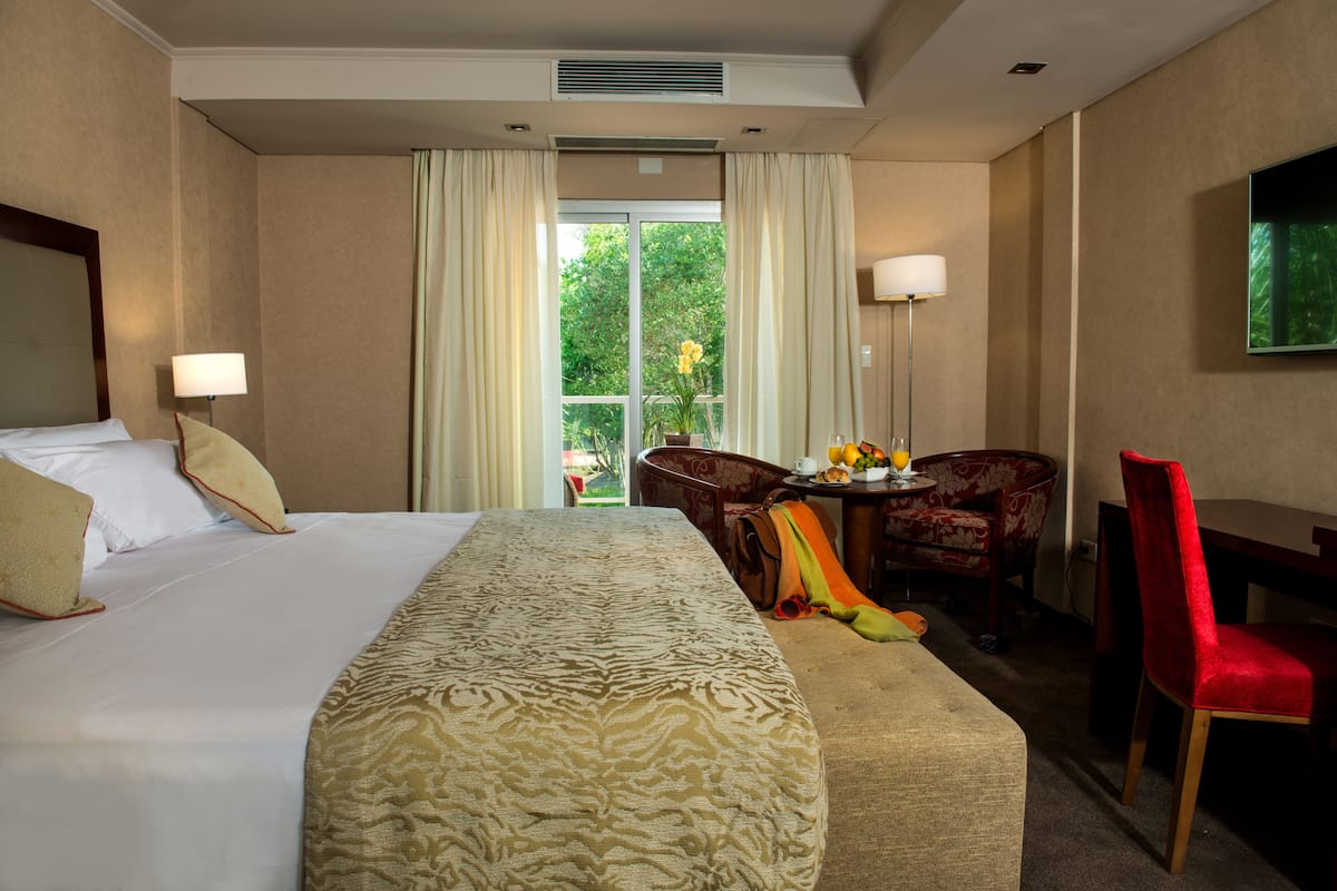 Quarto de hotel com uma cama grande, uma escrivaninha com cadeira, uma mesa com um vaso de flores e uma janela com cortinas.