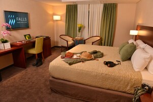 Quarto de hotel com cama, escrivaninha, cadeiras e televisão exibindo o nome do hotel.