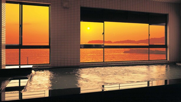 Quarto com grandes janelas voltadas para um corpo de água ao pôr do sol, parede com azulejos e área de piscina.