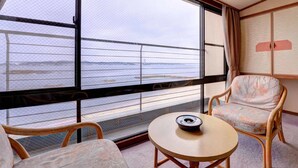 Quarto com uma grande janela com vista para um corpo d'água, uma mesa redonda com um objeto preto e duas cadeiras de vime com estofado floral.