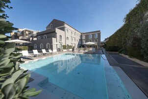 Piscina com espreguiçadeiras e guarda-sóis, rodeada por um edifício com várias varandas e um sebedeiro.