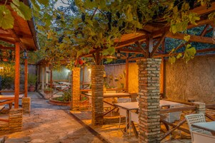 Uma área de pátio ao ar livre com piso de pedra, mesas e cadeiras de madeira e uma pérgola coberta por parras de uva.