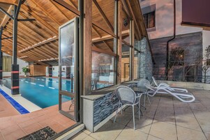 Piscina interior com parede de vidro, área de estar coberta com assentos e parede de pedra.