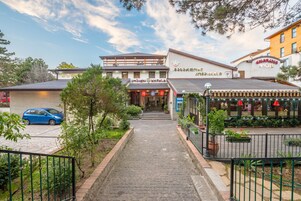 Entrada de hotel com caminho pavimentado até a porta, cercada por vegetação e um muro.