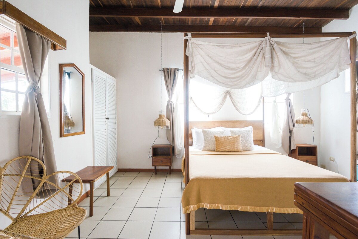 Um quarto com cama, mesas de cabeceira, uma cadeira e uma janela com cortinas.