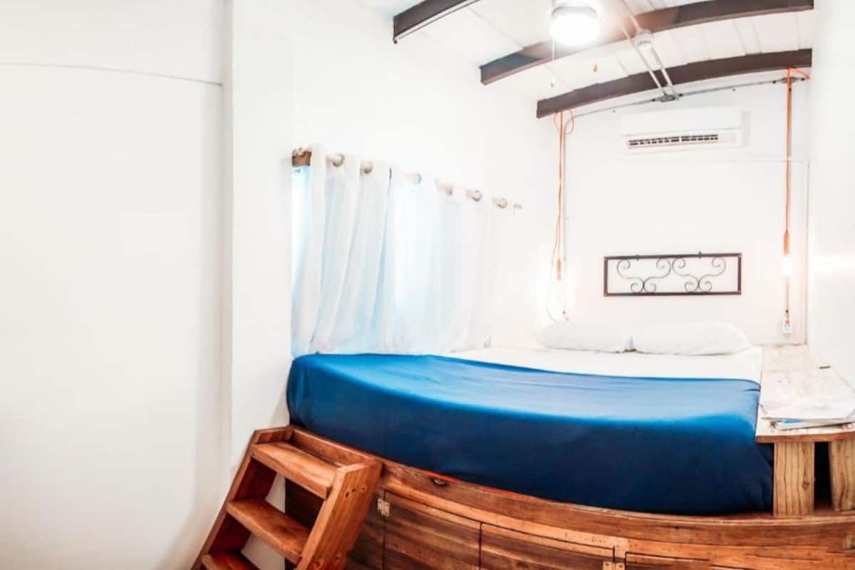 Quarto com cama, um banco de madeira, parede branca, luz no teto e um detalhe decorativo em metal.