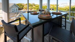 Uma área de refeições ao ar livre coberta, com cadeiras de vime e uma mesa de vidro, com vista para uma paisagem deslumbrante.
