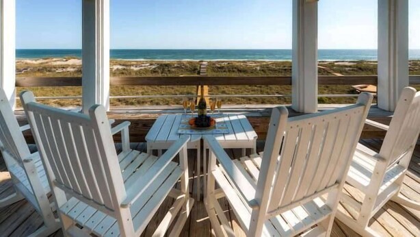 Um deck de madeira com cadeiras e mesa brancas, com vista para uma praia e o oceano ao fundo.