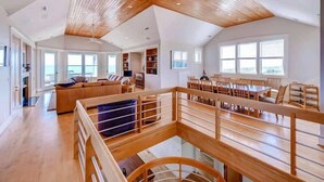 Uma sala de estar ampla com teto alto, vigas de madeira, área de jantar com mesa comprida e vista para o mar através de grandes janelas.