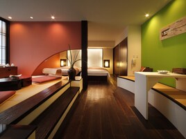 Quarto de hotel moderno com área de estar curvada, uma cama, uma mesa de trabalho e uma parede verde de destaque.