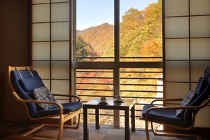 Quarto com duas cadeiras azuis, uma mesa pequena e uma janela grande com vista para árvores e colinas de outono.
