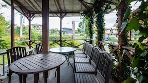 Uma área de estar coberta ao ar livre, com mesas e bancos de madeira, rodeada por vegetação e com vista para uma área residencial.
