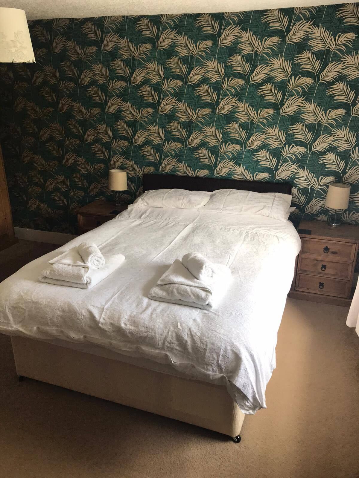 Cama bem arrumada com lençóis e toalhas brancas, papel de parede verde escuro estampado e duas mesas de cabeceira com abajures.