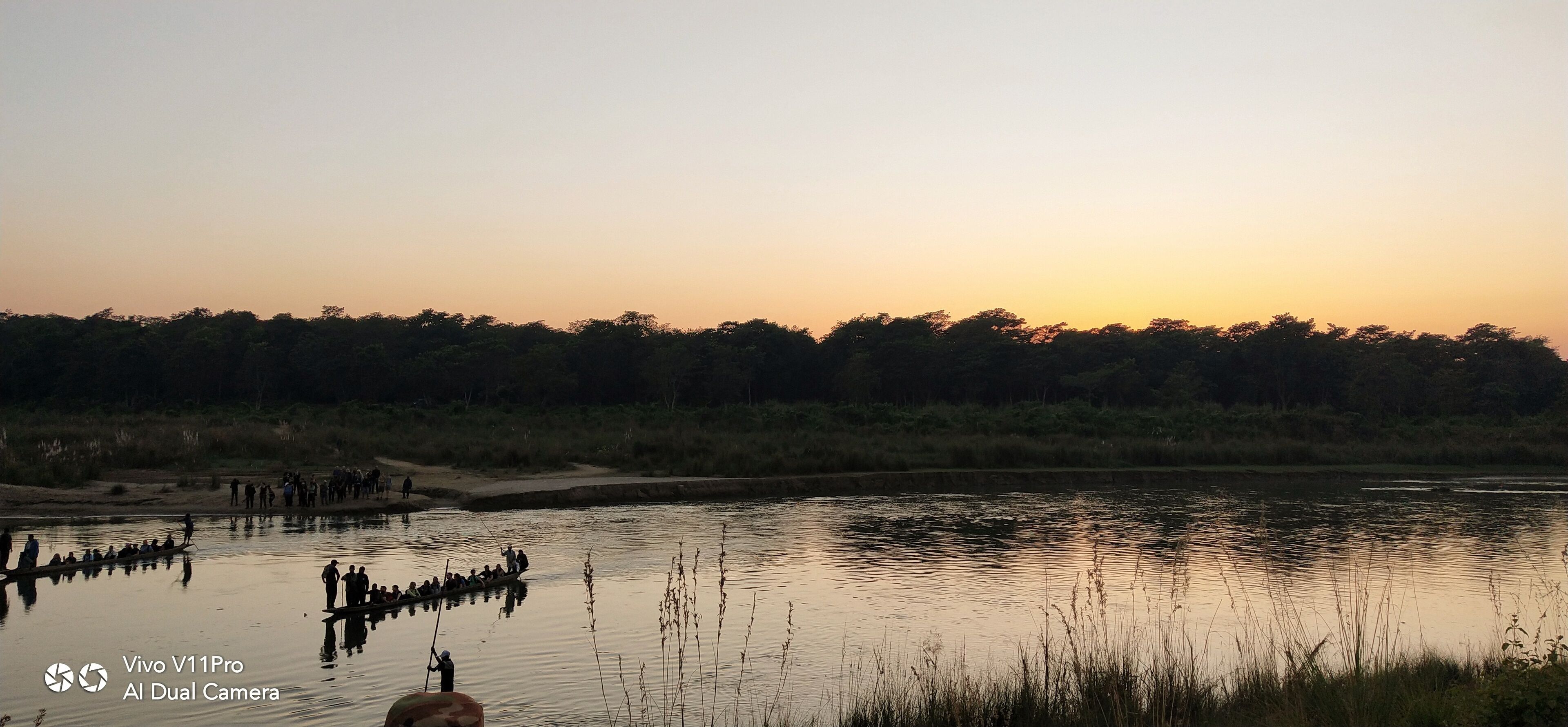 Um rio calmo com pessoas em canoas e barcos, cercado por árvores densas e um céu claro.