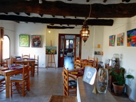 Uma sala de jantar com mesas e cadeiras de madeira, um quadro na parede e um candeeiro pendente.