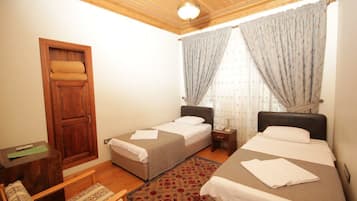 Quarto com duas camas de solteiro, piso de madeira, porta de madeira, uma mesa pequena com um objeto verde e um tapete estampado.
