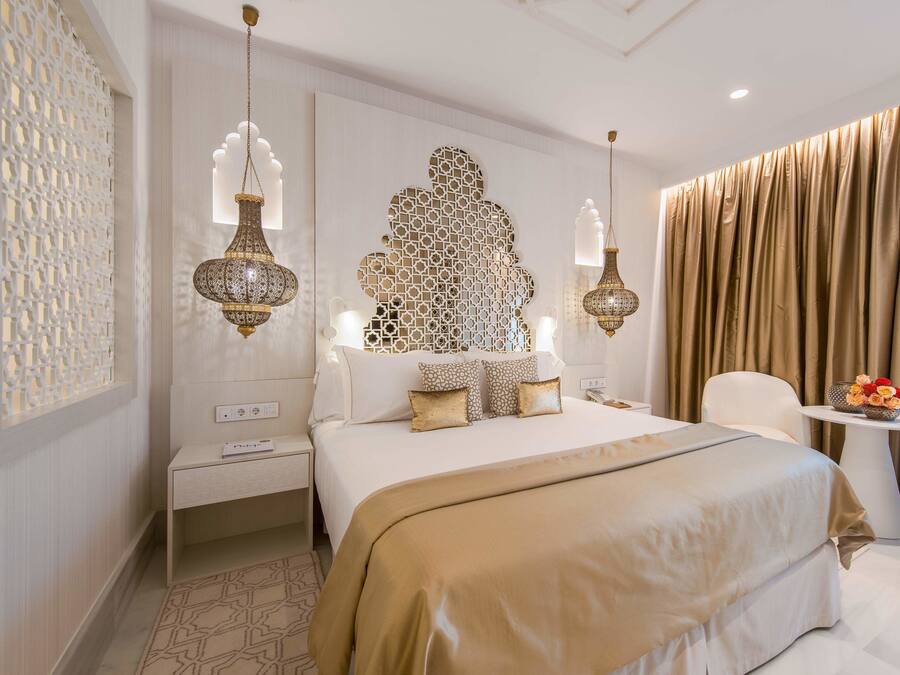 Quarto de hotel com uma cama grande, duas mesas de cabeceira, uma parede decorativa, um candelabro, uma mesa redonda com um vaso e uma cadeira.