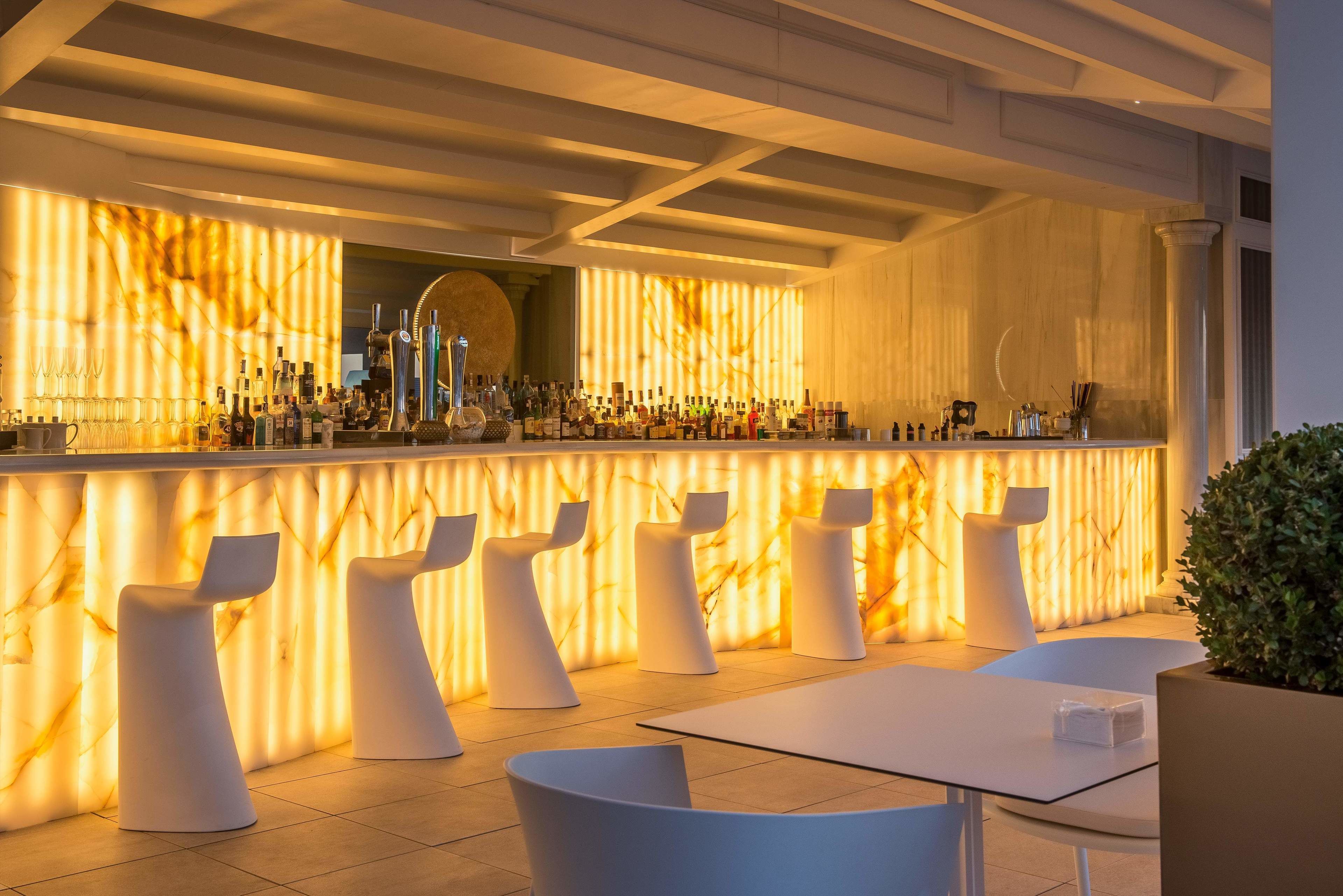 Uma área de bar com parede de mármore iluminada, banquetas brancas e diversos frascos e copos.