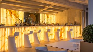 Uma área de bar com parede de mármore iluminada, banquetas brancas e diversos frascos e copos.