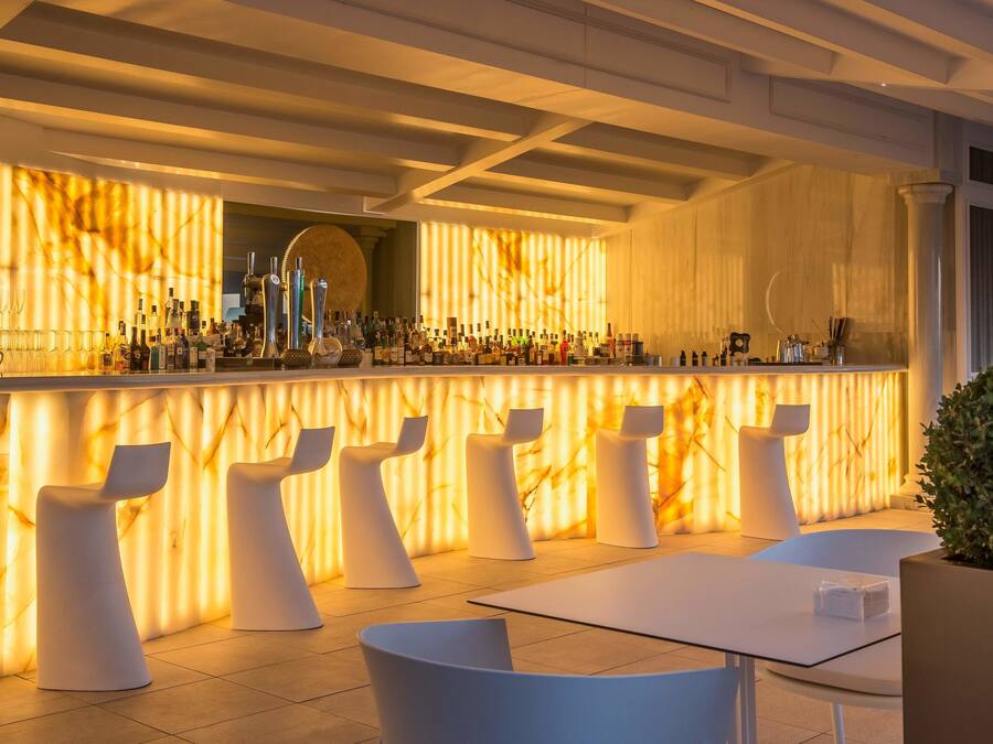 Uma área de bar com parede de mármore iluminada, banquetas brancas e diversos frascos e copos.