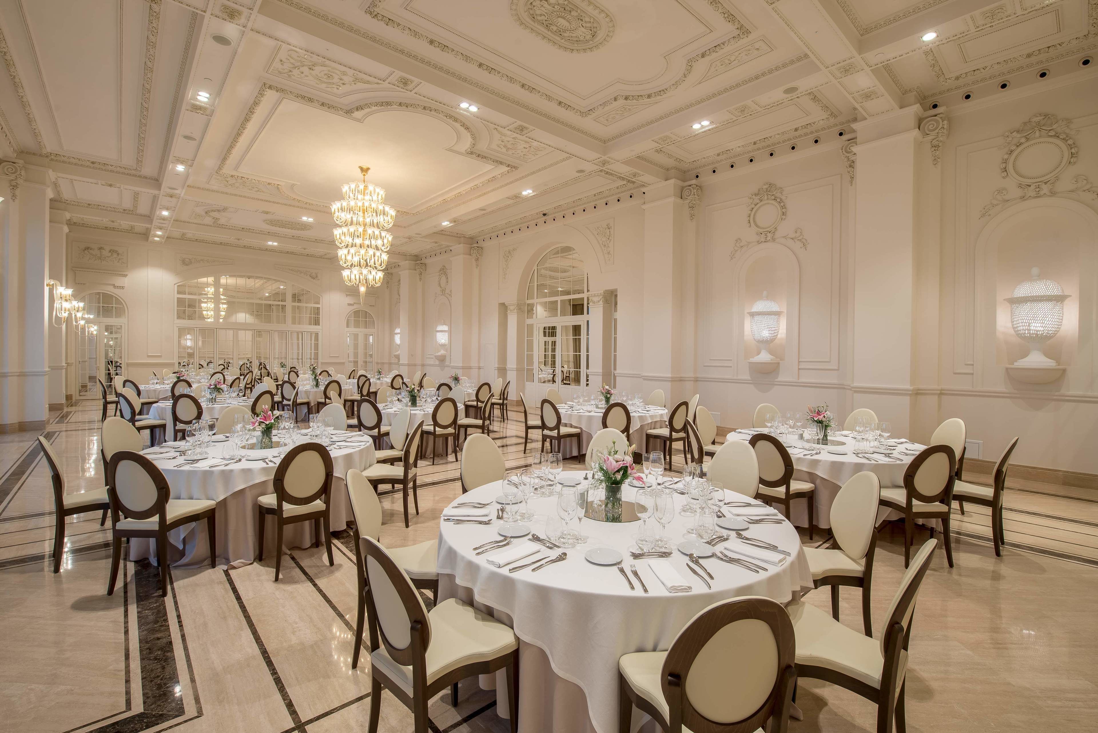 Uma sala de banquetes imponente com várias mesas redondas preparadas para refeições, lustres elegantes e detalhes arquitetônicos clássicos.