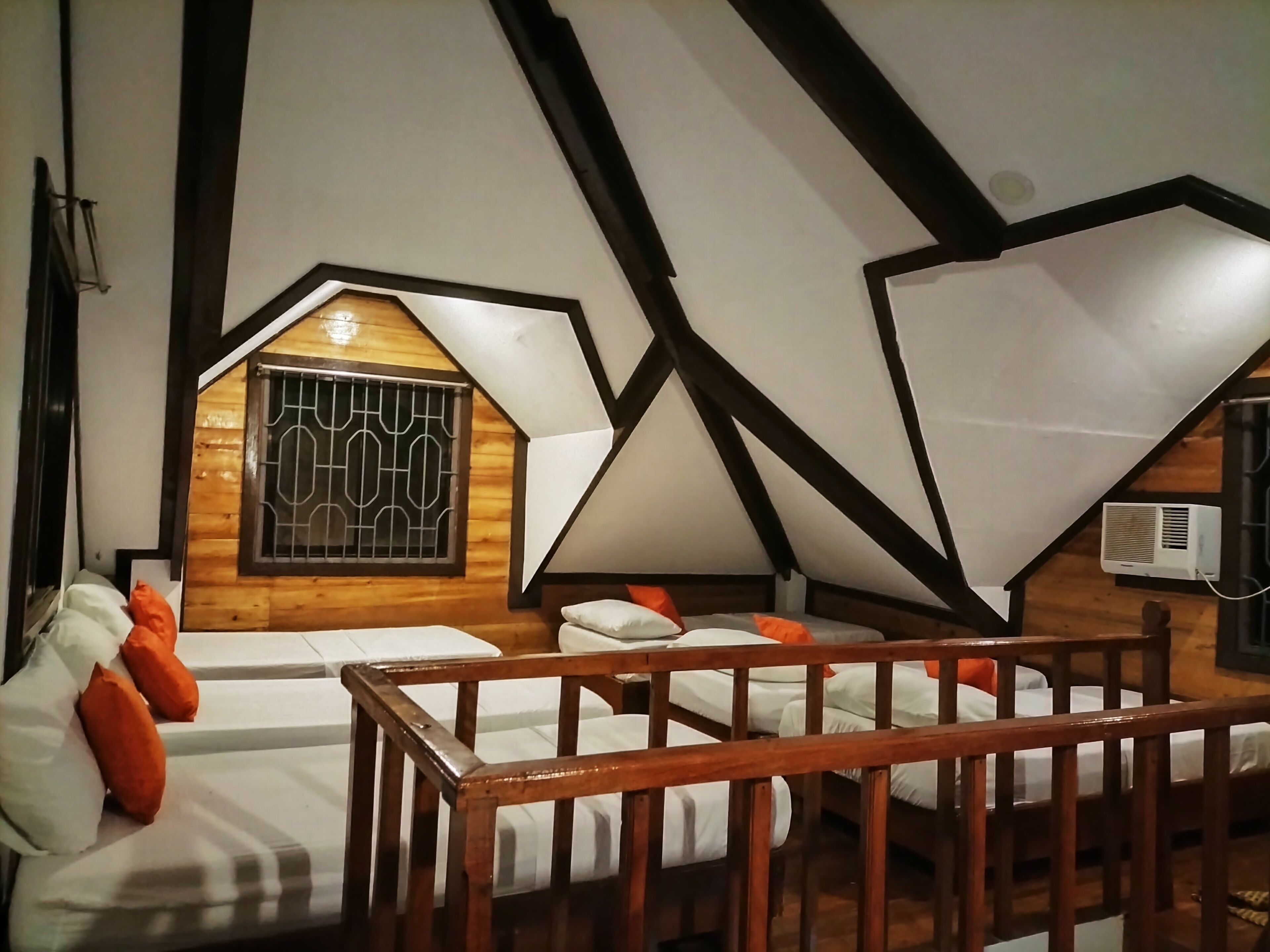 Quarto com teto inclinado, vigas de madeira e janela com grades decorativas. Há camas com roupa de cama branca e laranja.