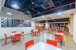 Uma sala de jantar moderna com cadeiras vermelhas e mesas brancas, uma cozinha ao fundo e uma grande obra de arte abstrata na parede.