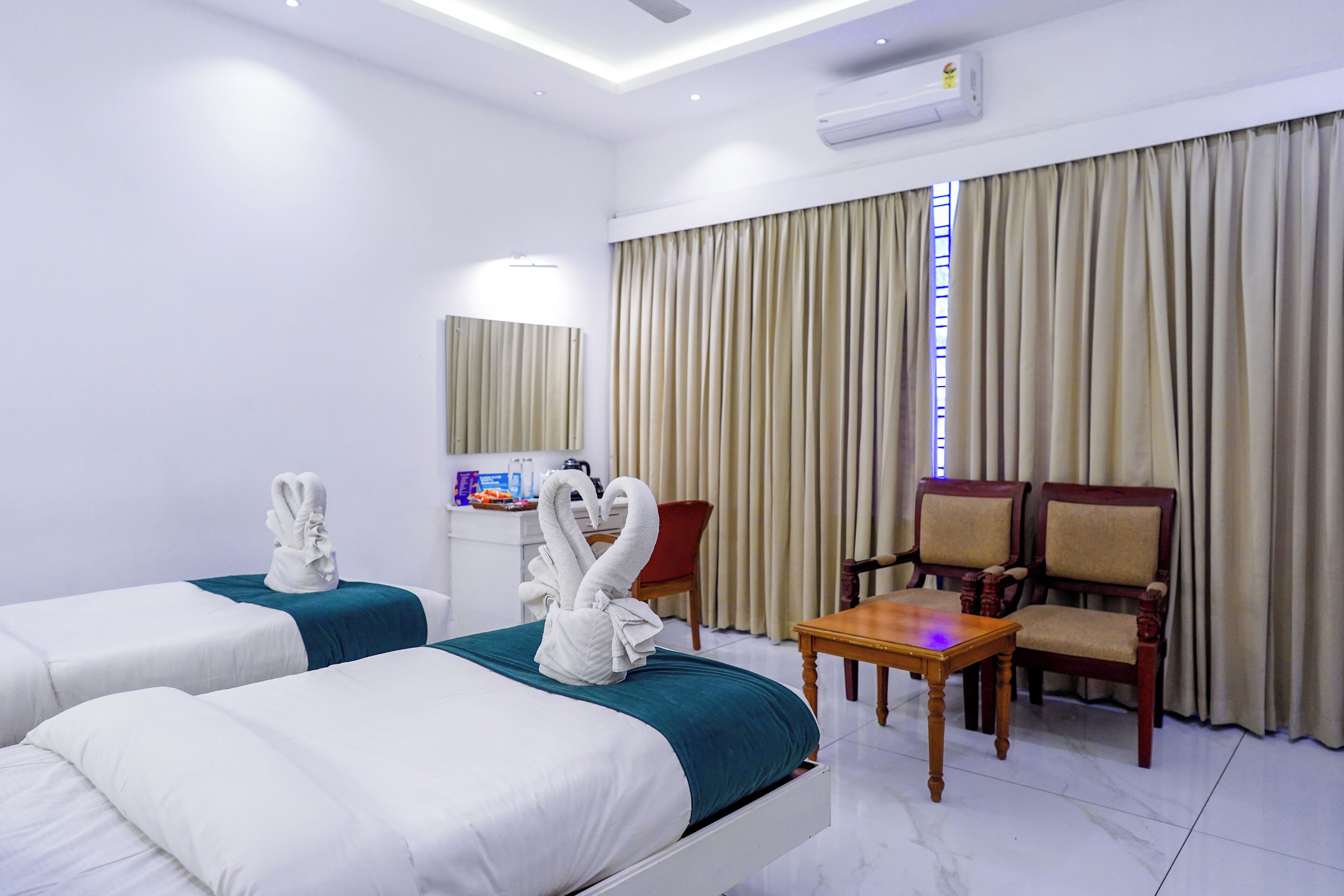 Quarto de hotel com duas camas, uma mesa de madeira, duas cadeiras, uma escrivaninha e uma janela com cortinas.